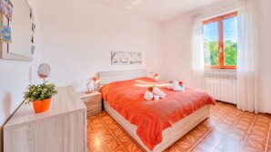 Quarto com cama, mesa de cabeceira, planta em vaso, janela com cortinas e espelho fixado na parede.