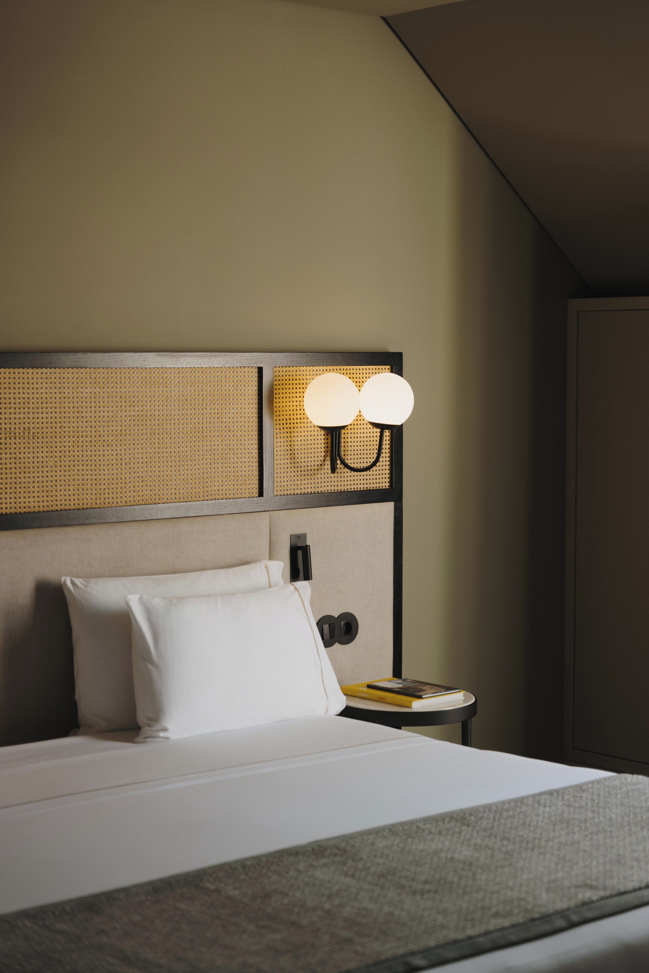 Uma cama bem arrumada com lençóis brancos, cabeceira com painel trançado e mesa de cabeceira com um livro e uma luminária.