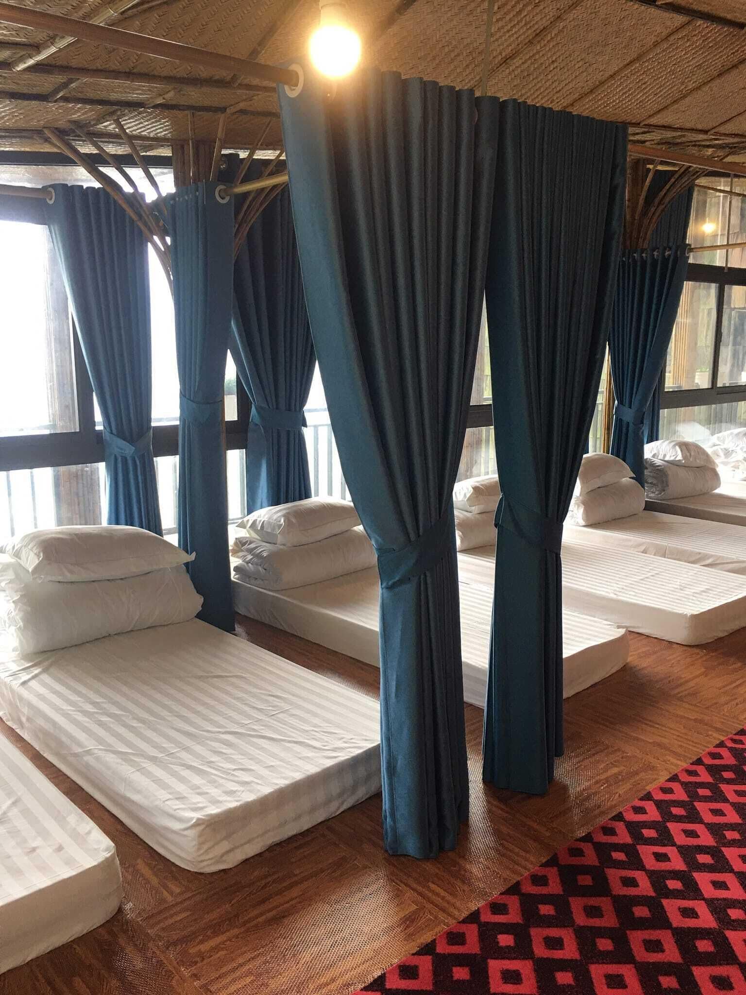 Quarto com quatro camas individuais, cada uma com roupa de cama e almofadas brancas. O quarto possui piso de madeira e tapete estampado. Cortinas azuis estão fechadas sobre as janelas, e uma luminária é visível no teto.