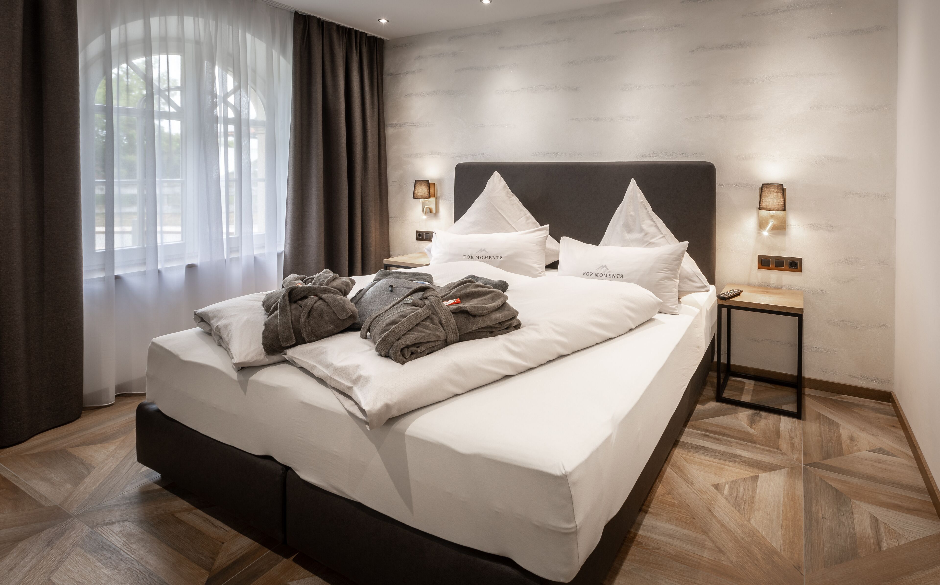 Cama bem arrumada com lençóis brancos e roupões cinzas, piso de madeira e janela com cortinas leves.