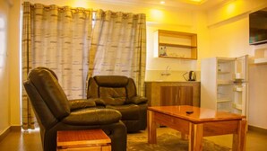 Sala de estar com sofá de couro, mesa de centro de madeira e uma mesinha lateral. Há uma área de cozinha com pia e armários, e uma televisão fixada na parede.