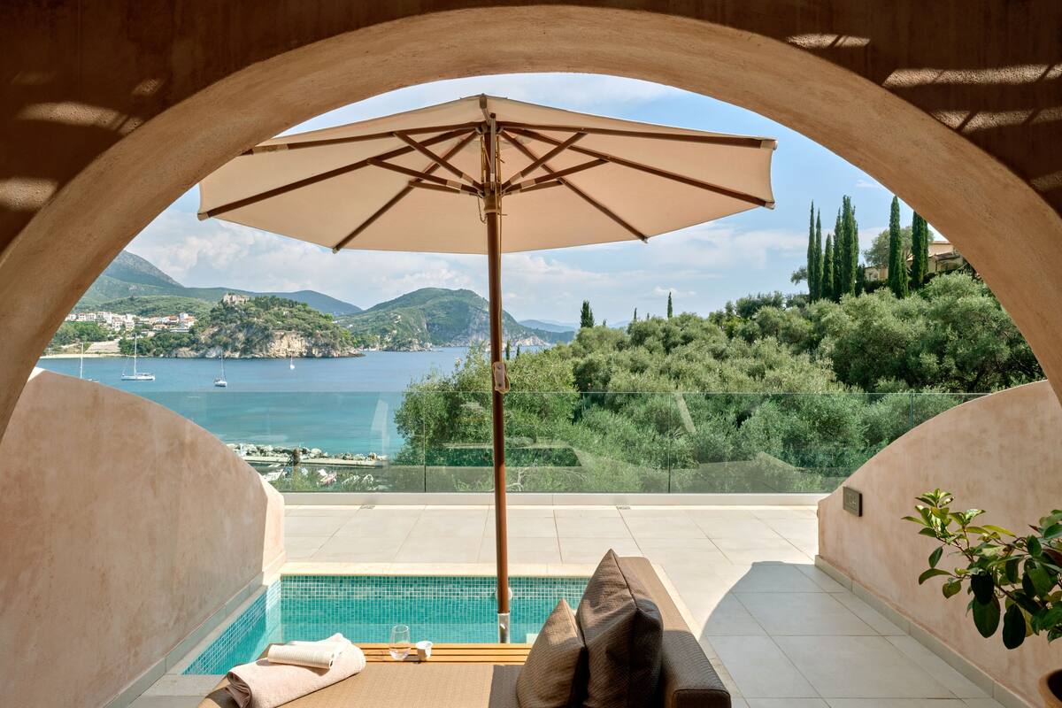 Uma varanda com piscina, um sofá e um guarda-sol, com vista para o mar e montanhas.