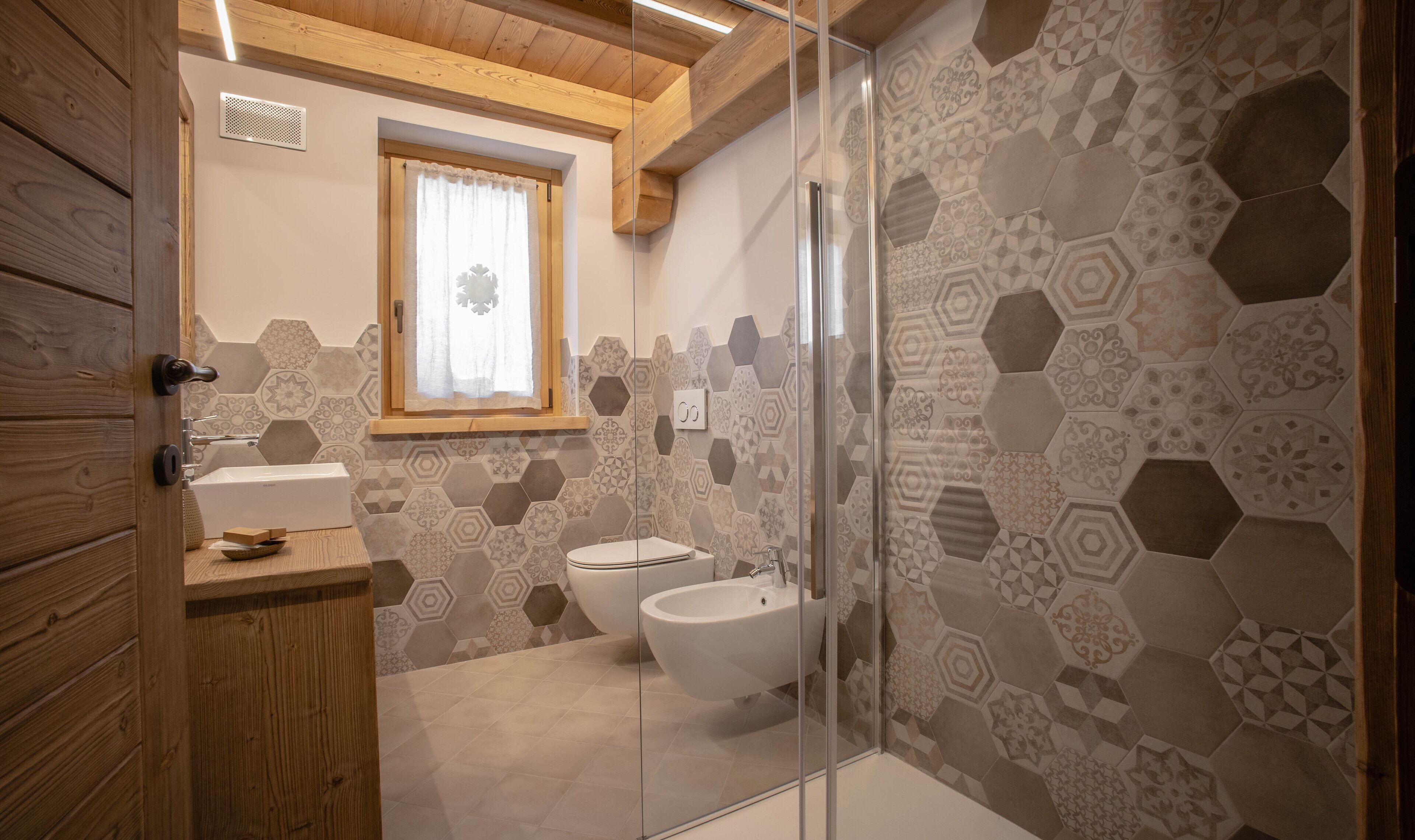 Uma casa de banho moderna com cabine de duche em vidro, sanita e bidé. As paredes são decoradas com azulejos com padrão hexagonal.