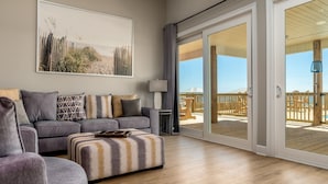 Sala de estar moderna com sofá cinza, banquinho estampado e uma grande janela com vista para um deck de madeira e praia.