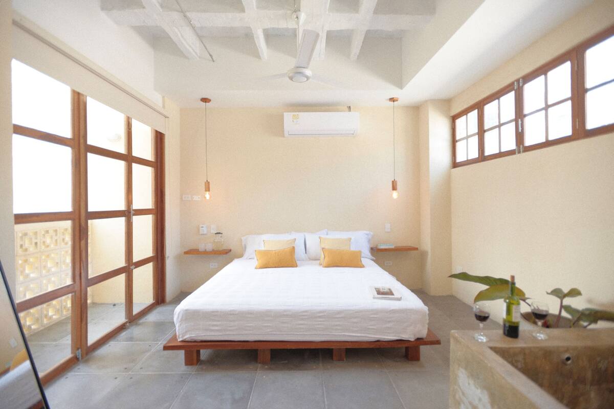 Um quarto espaçoso com uma cama grande, cabeceira de madeira e um ventilador de teto. Há duas mesas de cabeceira com luminárias, uma janela com vista e um aparelho de ar condicionado na parede.