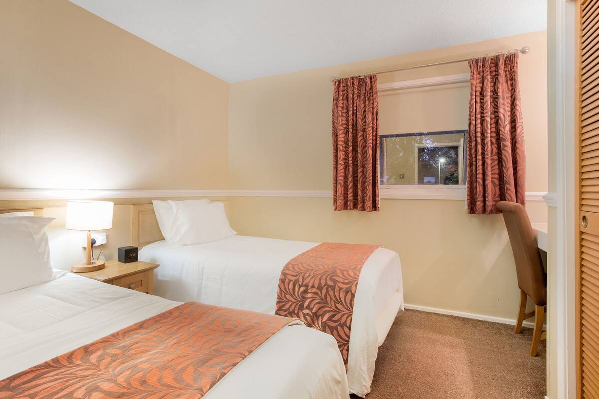 Quarto de hotel com duas camas, uma mesa de cabeceira com abajur, uma janela com cortinas e uma cadeira.