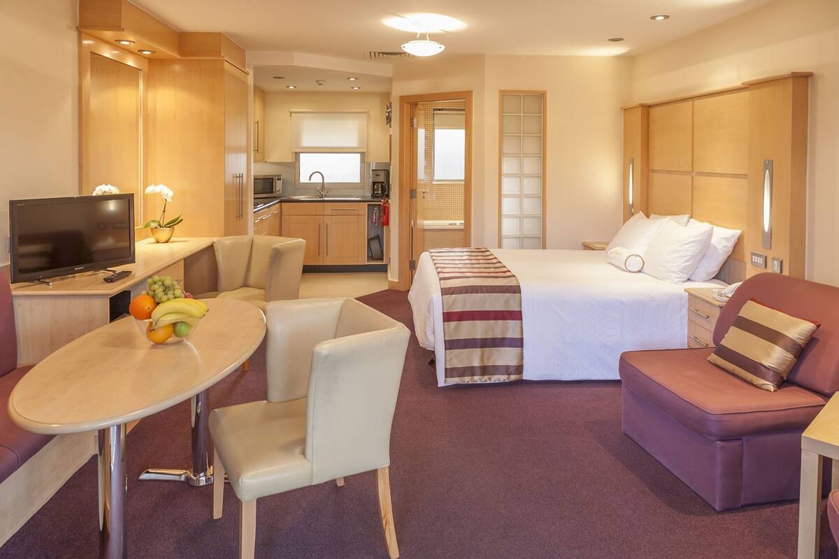 Quarto de hotel com cama, sofá, televisão, uma pequena mesa com frutas e cozinha compacta.
