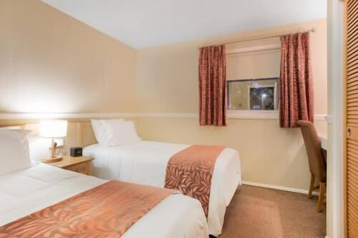 Quarto de hotel com duas camas, uma janela com cortinas, uma mesinha de cabeceira com abajur e uma cadeira.