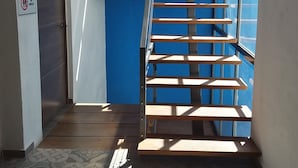 Escadaria com corrimão de metal e degraus de madeira, levando a um quarto com ladrilhos estampados e parede azul.