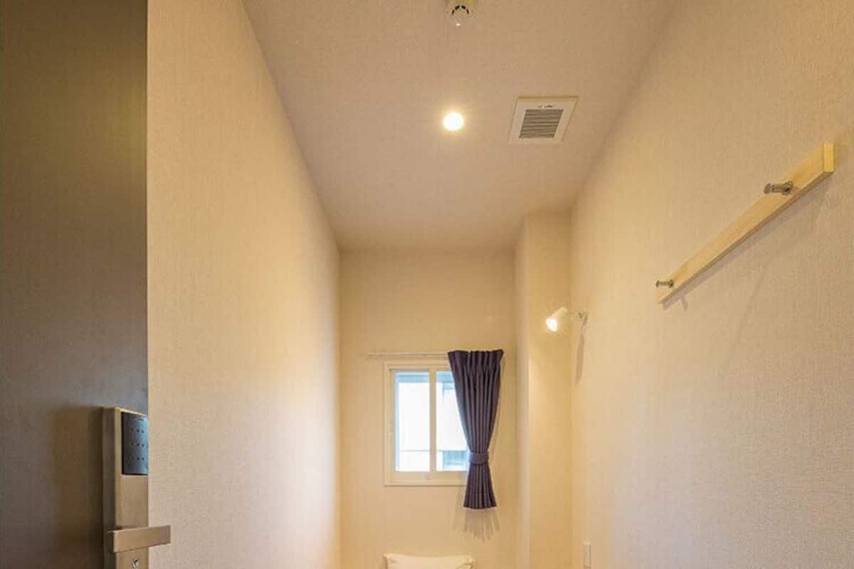 Quarto compacto com cama, uma pequena janela com cortinas, mesa de cabeceira e luminária fixada na parede.