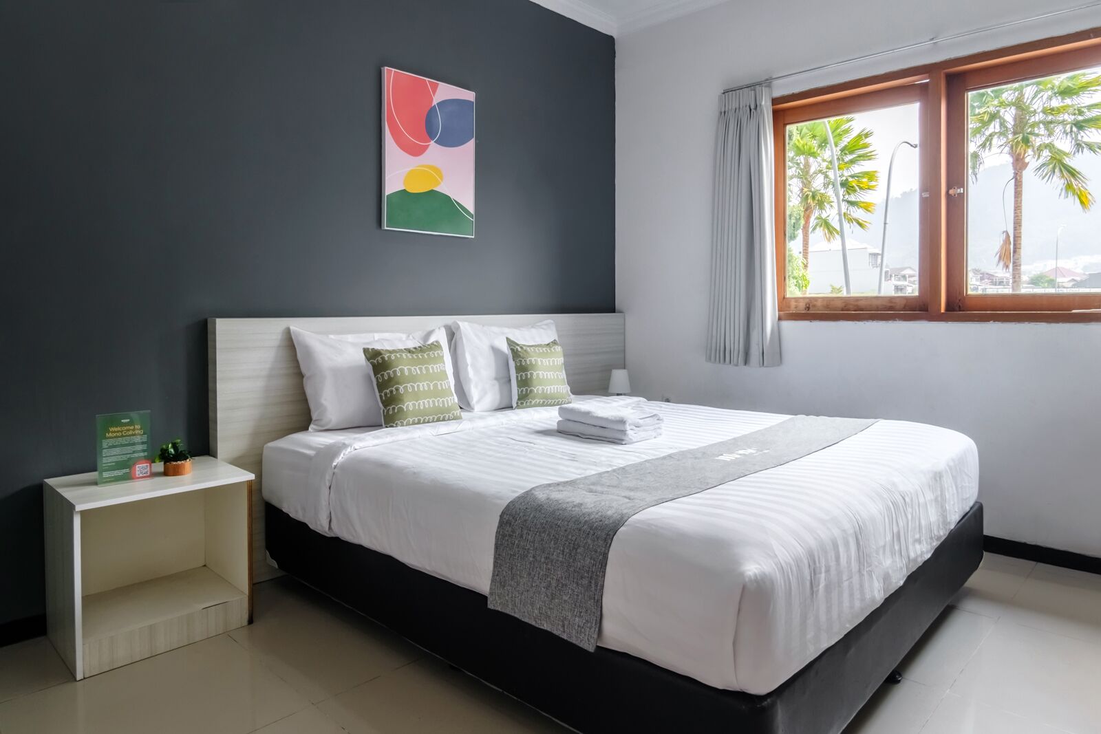 Quarto de hotel moderno com uma cama grande, uma mesinha de cabeceira, um quadro e uma janela com vista para palmeiras.