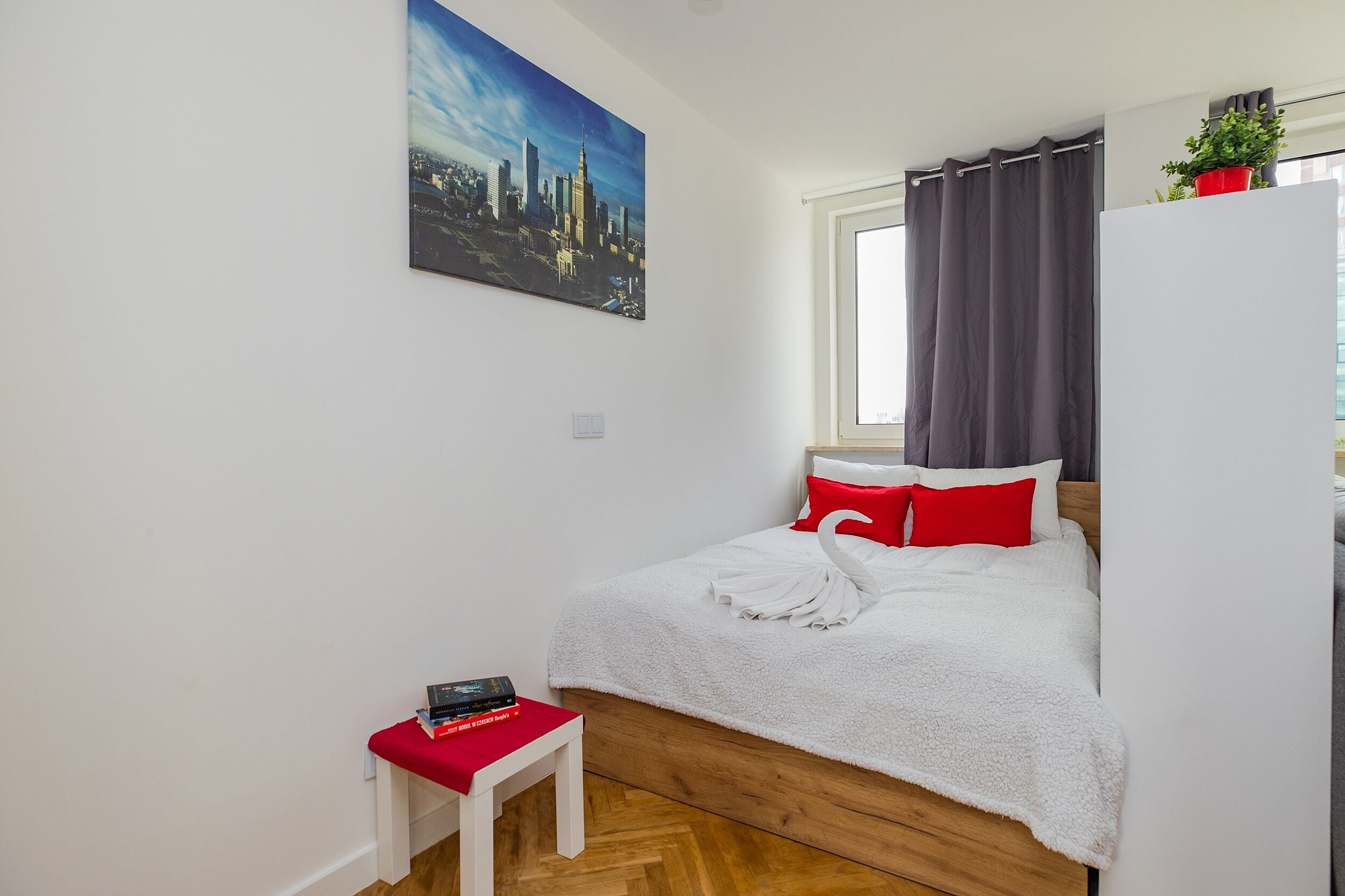 Quarto com cama, uma mesinha com livros, janela com cortinas e uma pintura da paisagem urbana na parede.