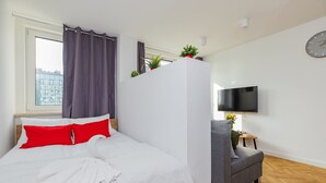 Quarto de hotel moderno com uma cama, almofadas vermelhas, uma poltrona cinza, uma TV e uma janela com cortinas.