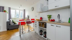 Uma cozinha moderna com armários brancos, bancada de madeira e banquetas vermelhas. Há uma janela com cortinas, um sofá e um relógio na parede.