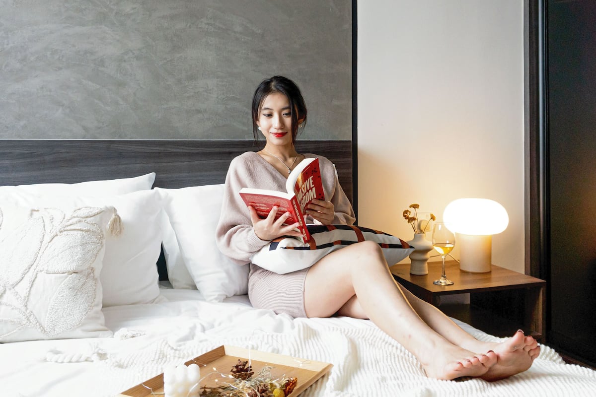 Uma pessoa sentada na cama com um livro, uma mesinha de cabeceira com uma luminária e uma janela.
