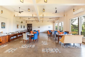 Ampla sala de jantar com várias mesas e cadeiras, ventiladores de teto e serviço de buffet.