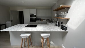 Uma cozinha moderna com bancada de mármore, dois bancos de bar brancos, fornos embutidos, pia e adega a vinho.