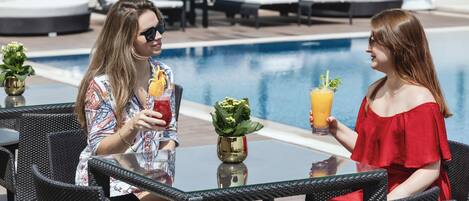 Duas mulheres sentadas à mesa perto de uma piscina, com espreguiçadeiras e uma cama ao fundo.