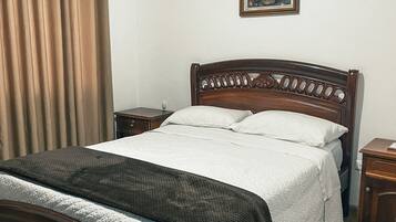 Quarto com cama de madeira, roupa de cama branca, mesa de cabeceira e um quadro floral emoldurado na parede, além de uma janela com cortinas.