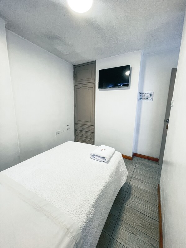 Quarto de hotel com uma cama de solteiro, televisão presa à parede e porta que dá para outro quarto.