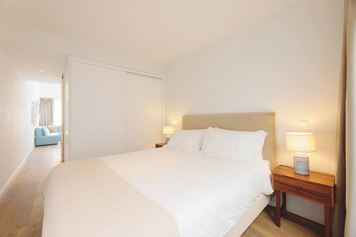 Uma cama bem arrumada com roupa de cama branca, uma mesa de cabeceira com abajur e vista para um sofá azul no quarto vizinho.