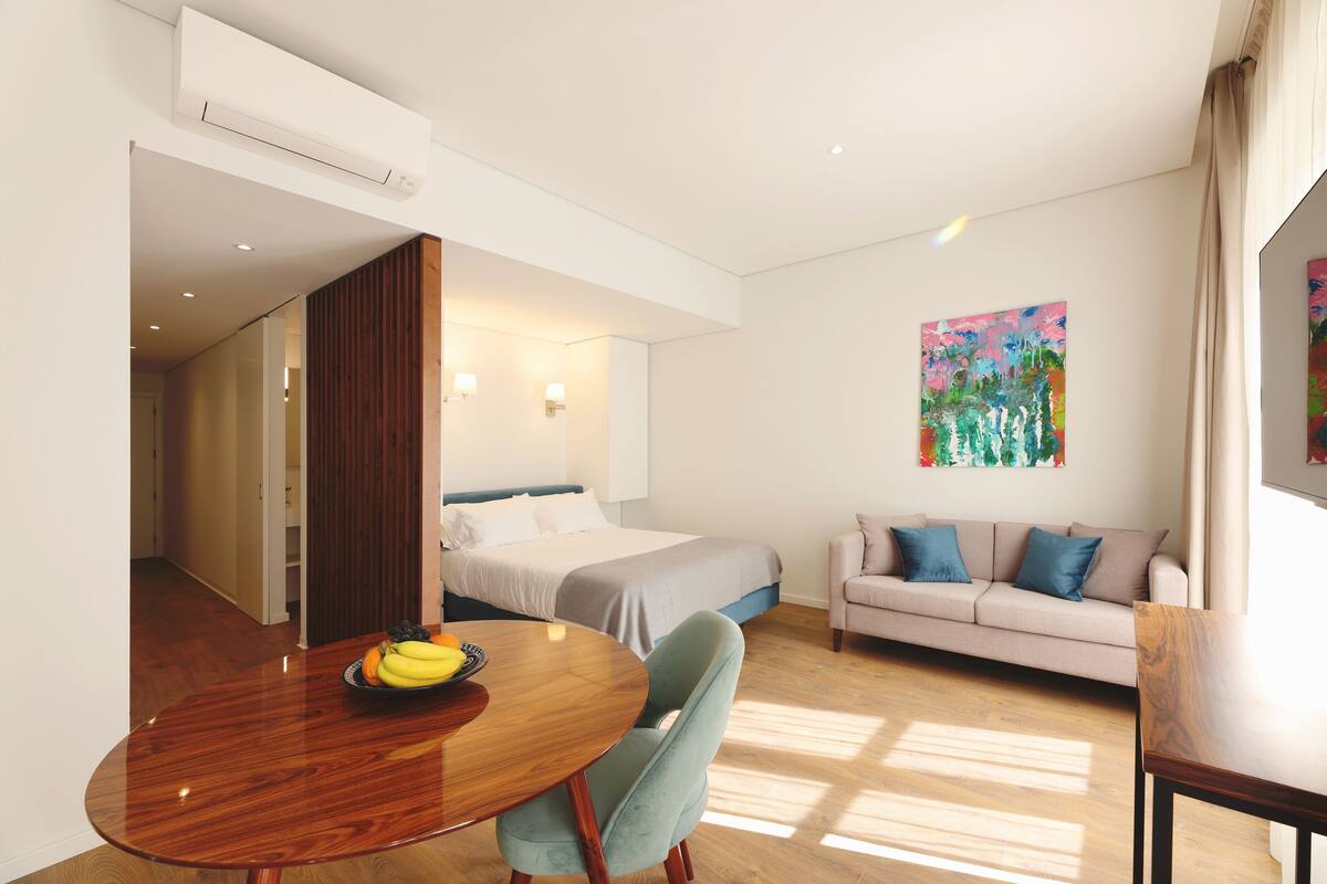 Quarto de hotel com uma cama, um sofá, uma mesa de jantar com frutas e um quadro na parede.