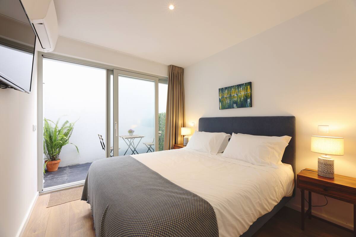 Quarto de hotel moderno com uma cama grande, uma mesinha, uma planta em vaso e vista para o exterior.