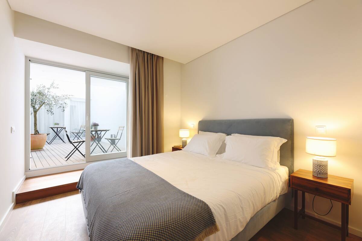 Quarto de hotel com uma cama grande, uma mesa de cabeceira com abajur, uma porta de vidro que desliza para área externa com mesa e cadeiras, e vista para uma árvore.
