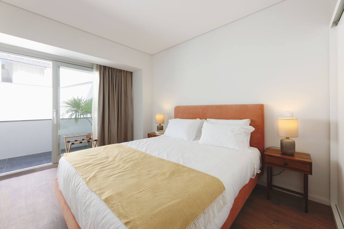 Quarto de hotel com uma cama, duas mesas de cabeceira com luminárias, uma porta de vidro deslizante para a varanda e uma pequena mesa com uma cadeira.