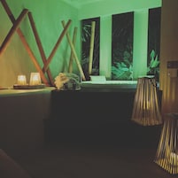 Quarto com design minimalista, apresentando galhos de bambu, uma pequena mesa com velas e vista para plantas tropicais pela janela.