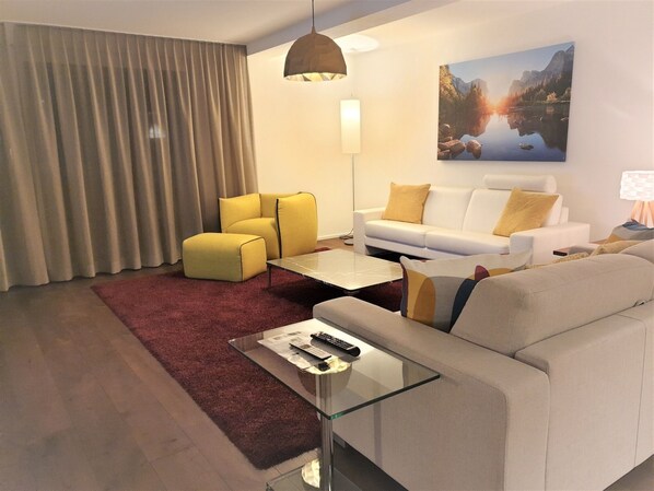 Uma sala de estar moderna com uma mesa de centro de vidro, um sofá, uma poltrona amarela e um sofá-cama branco.