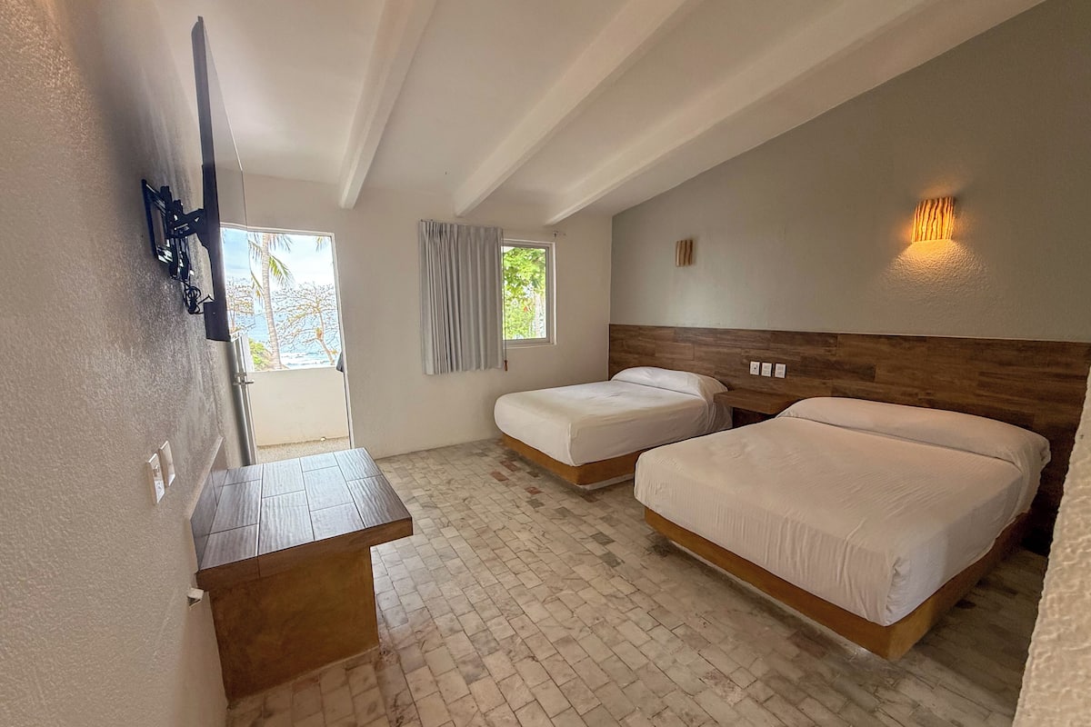 Quarto de hotel com duas camas, cabeceira de madeira, uma mesinha e televisão fixada na parede.