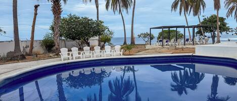 Piscina com cadeiras brancas e palmeiras, área de estar coberta e vista para o oceano.