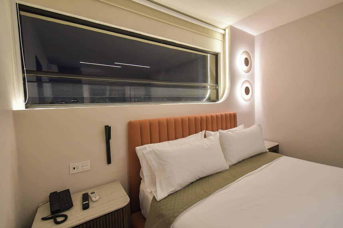 Quarto de hotel com uma janela grande, uma cama com roupa de cama branca, mesa de cabeceira com telefone e controle remoto, e luminária fixa na parede.