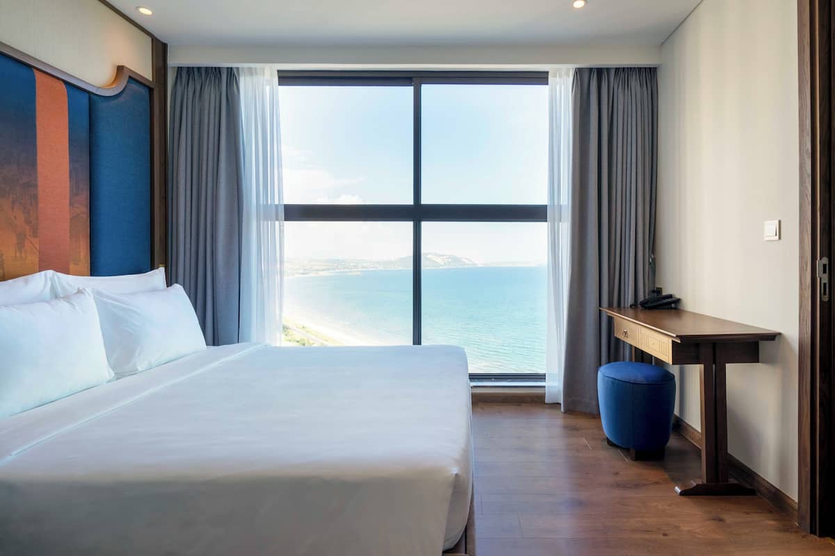 Quarto de hotel com cama, escrivaninha, cadeira e uma janela grande com vista para o mar.