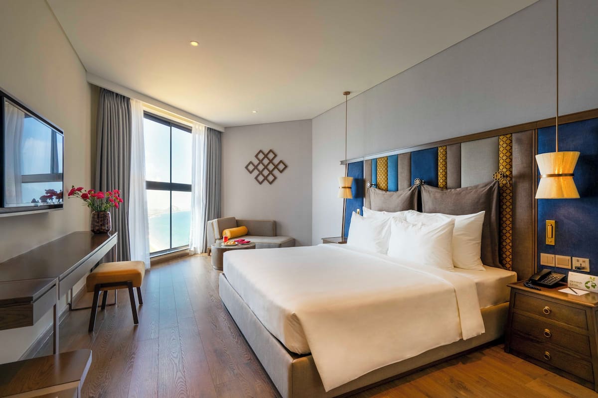 Quarto de hotel com uma cama grande, uma escrivaninha, uma cadeira, televisão e janela com cortinas.