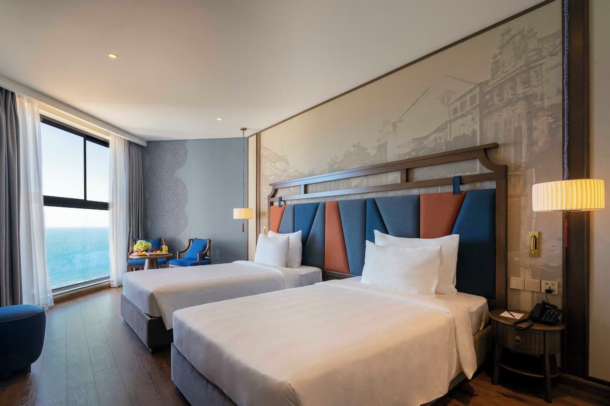 Quarto de hotel com duas camas, uma escrivaninha, uma cadeira, uma luminária e vista para o mar.