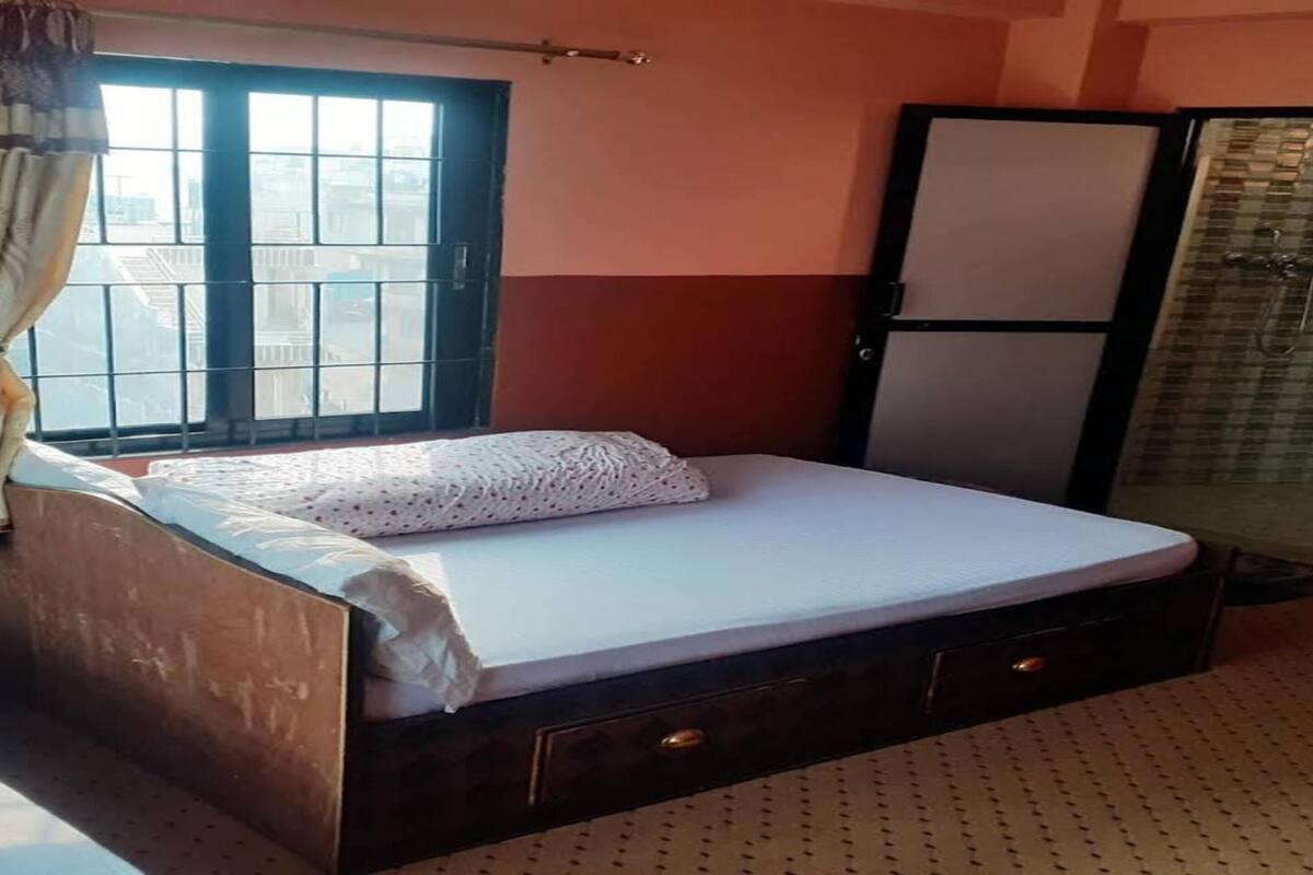 Uma cama de solteiro com colchão branco e almofada estampada, janela com cortinas e chuveiro com porta de vidro.