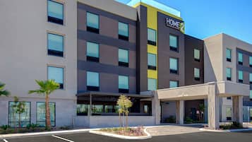 Um hotel moderno com a placa "HOME 2", apresentando uma construção em vários andares, grandes janelas e uma área de entrada coberta.