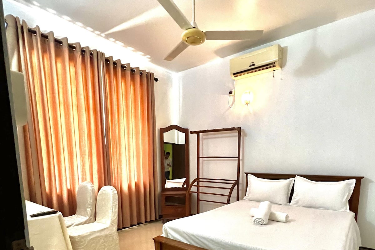 Quarto de hotel com cama, ventoinha no teto, cadeira, escrivaninha e espelho.
