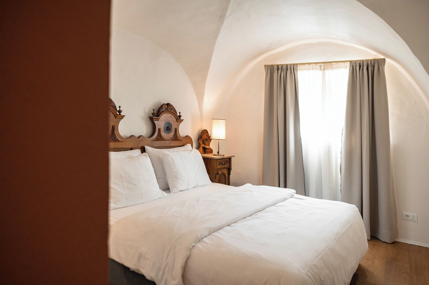 Quarto com uma cama grande, cabeceira de madeira, mesa de cabeceira com abajur e janela com cortinas.