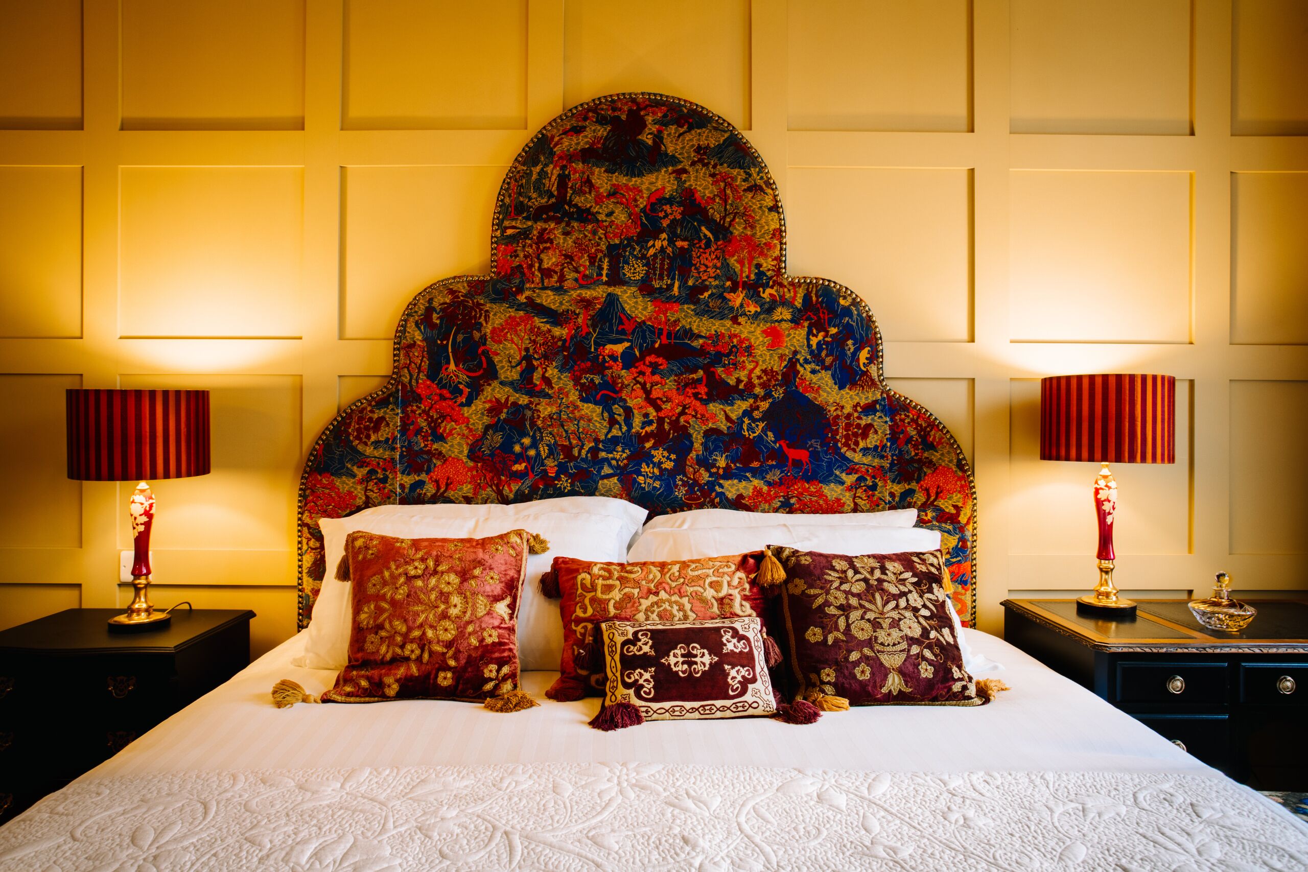 Cama com cabeceira elaborada, várias almofadas e luminárias laterais.