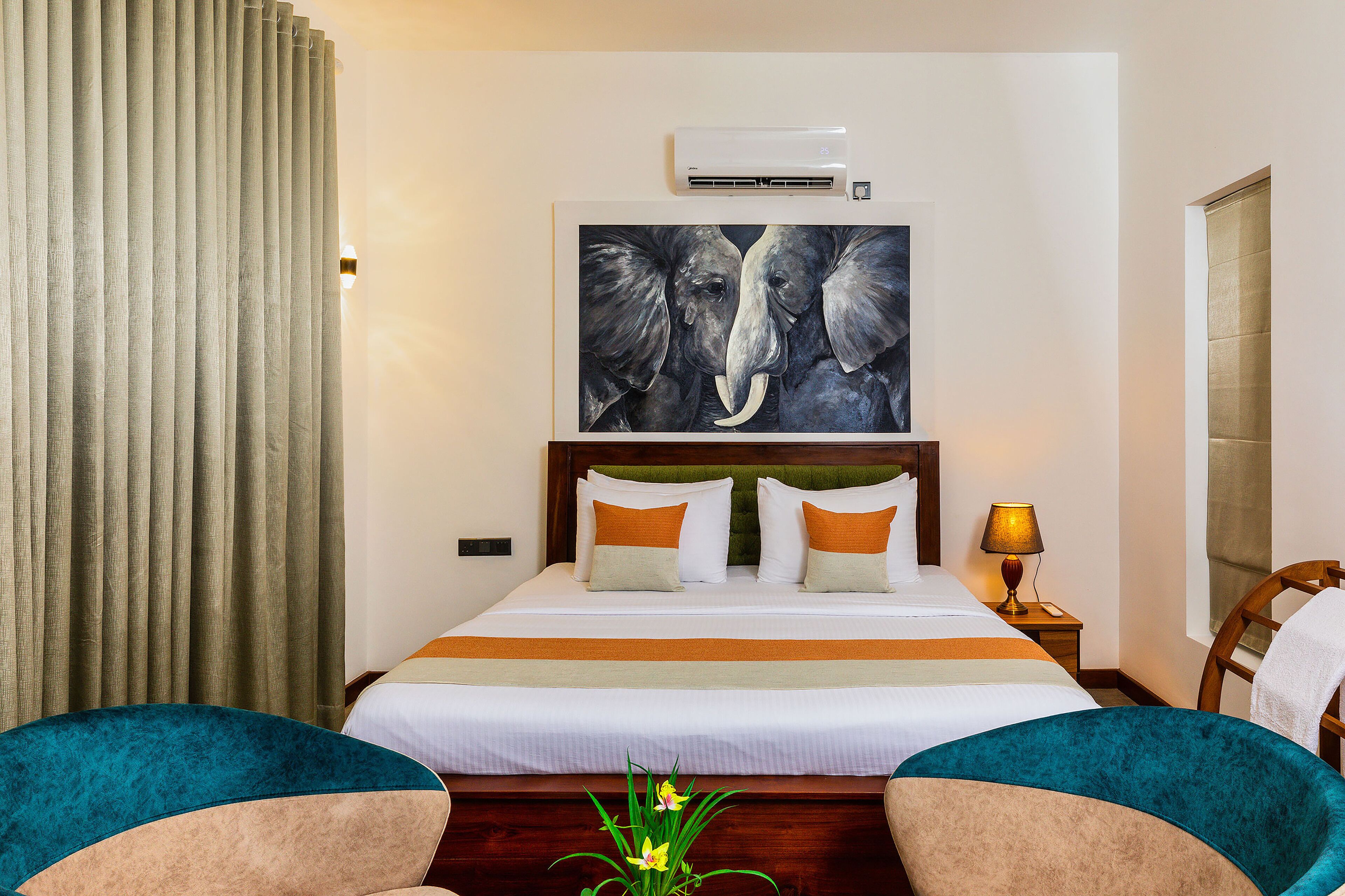 Quarto de hotel com uma cama grande, duas cadeiras, um quadro de elefantes, ar condicionado e uma luminária.