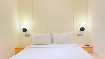 Cama bem arrumada com lençóis brancos num quarto com paredes em tom bege e teto inclinado. Há duas mesas de cabeceira com luminárias de cada lado da cama.