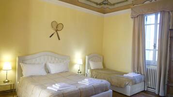 Quarto de hotel com duas camas, uma janela com cortinas, um radiador e uma raquete de ténis presa à parede.