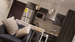 Uma sala de estar moderna com um sofá, mesa de centro e controles remotos. Uma cozinha com eletrodomésticos de aço inoxidável e uma área de jantar com mesa e cadeiras são visíveis ao fundo.