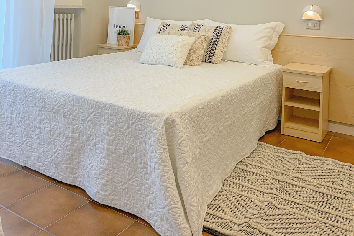 Uma cama bem arrumada com colcha estampada, uma mesa de cabeceira com abajur e um tapete no chão.