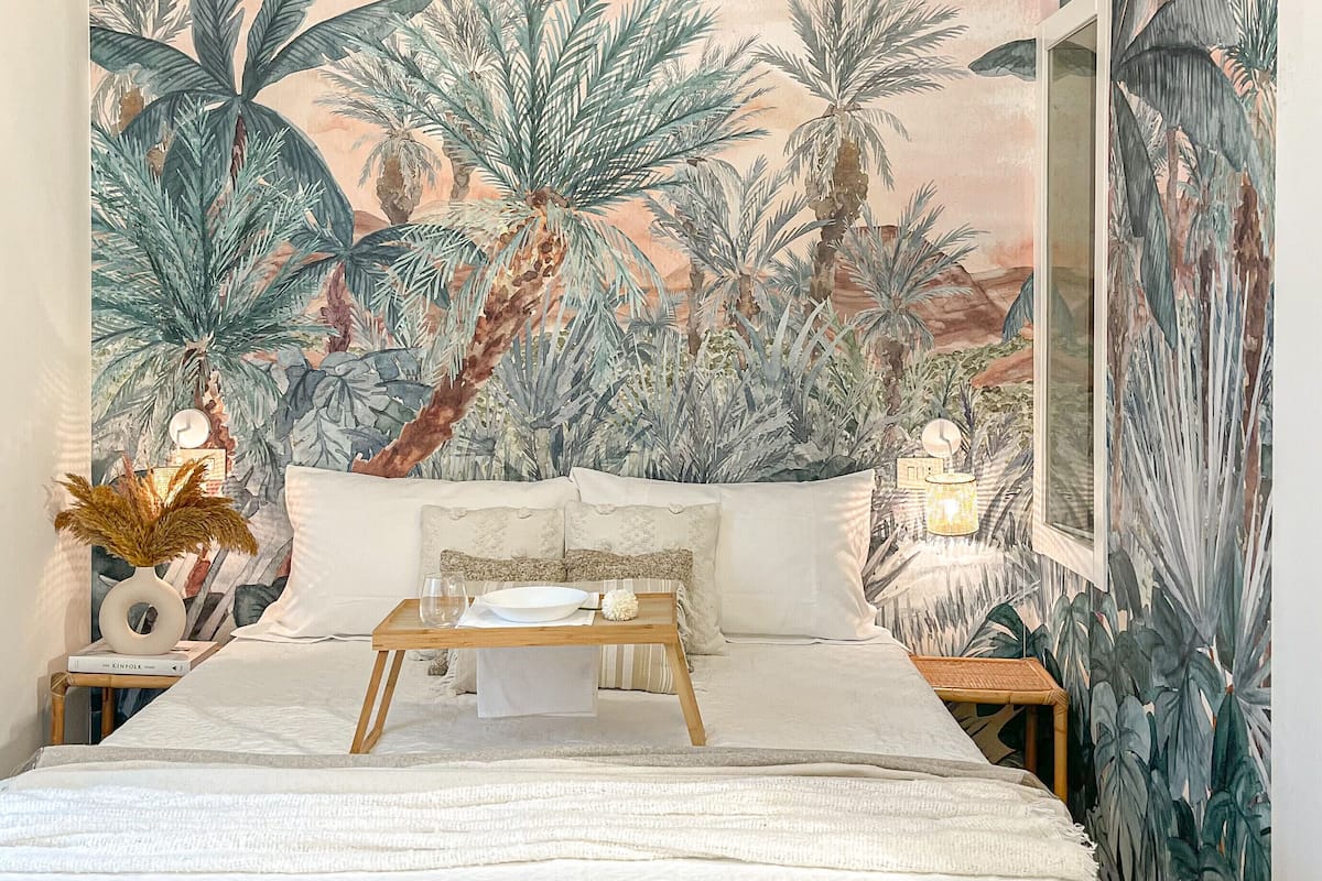 Quarto com um grande mural de folhagem tropical, cama com colcha texturizada branca e mesa de cabeceira com um vaso e livros.