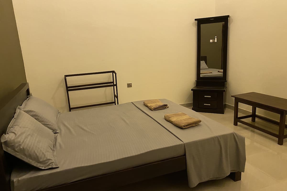 Cama bem arrumada com um edredom cinza e duas toalhas dobradas, uma mesinha de cabeceira de madeira com espelho, e um pequeno banco de madeira.