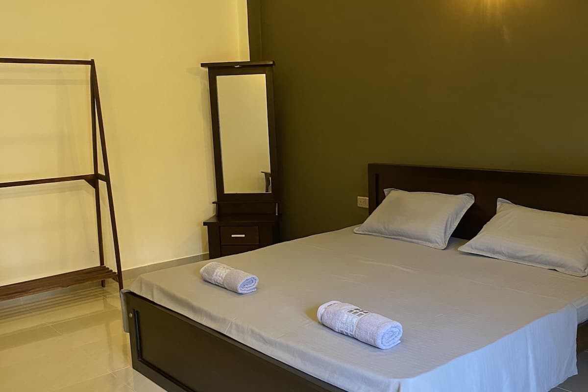 Cama bem arrumada com lençóis brancos e toalhas enroladas, mesa de cabeceira com espelho e luminária fixada na parede.